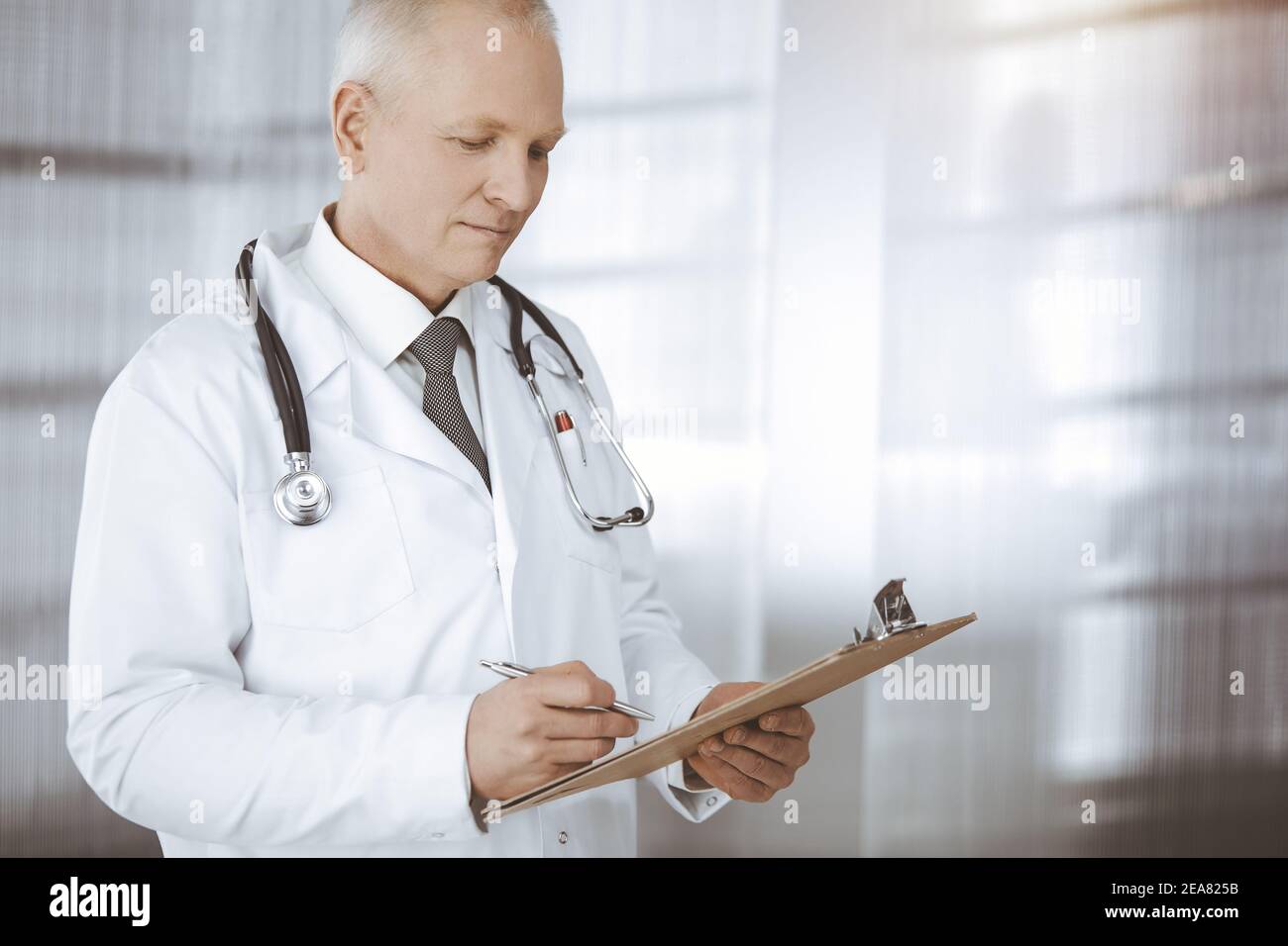 Elderly confident doctor standing straight and using clipboard in sunny ...