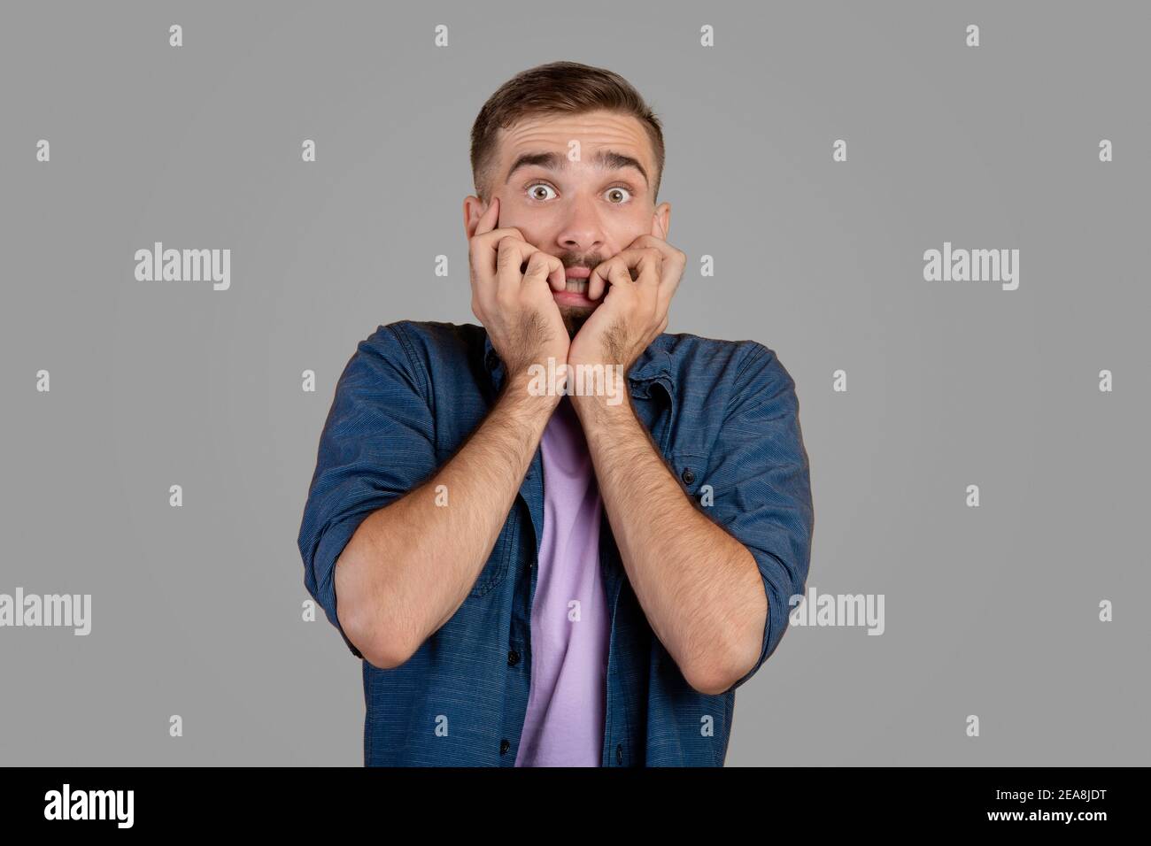 Scared Caucasian guy biting his nails in terror over grey studio ...