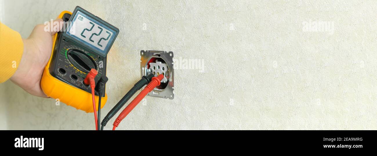 Male electrician checks the voltage of an outlet on a white wall with a ...