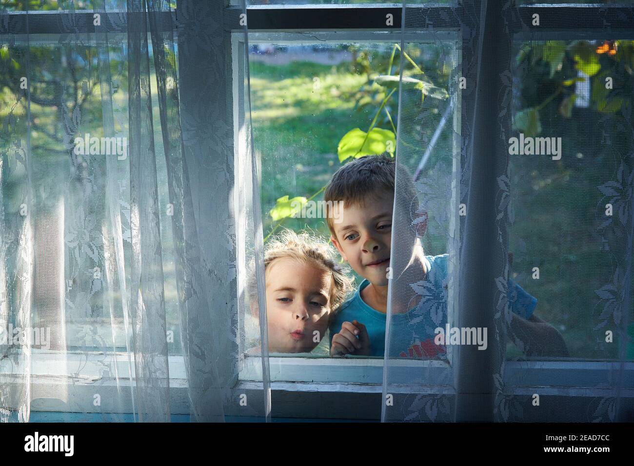 Two funny children are looking through the window inside the house ...