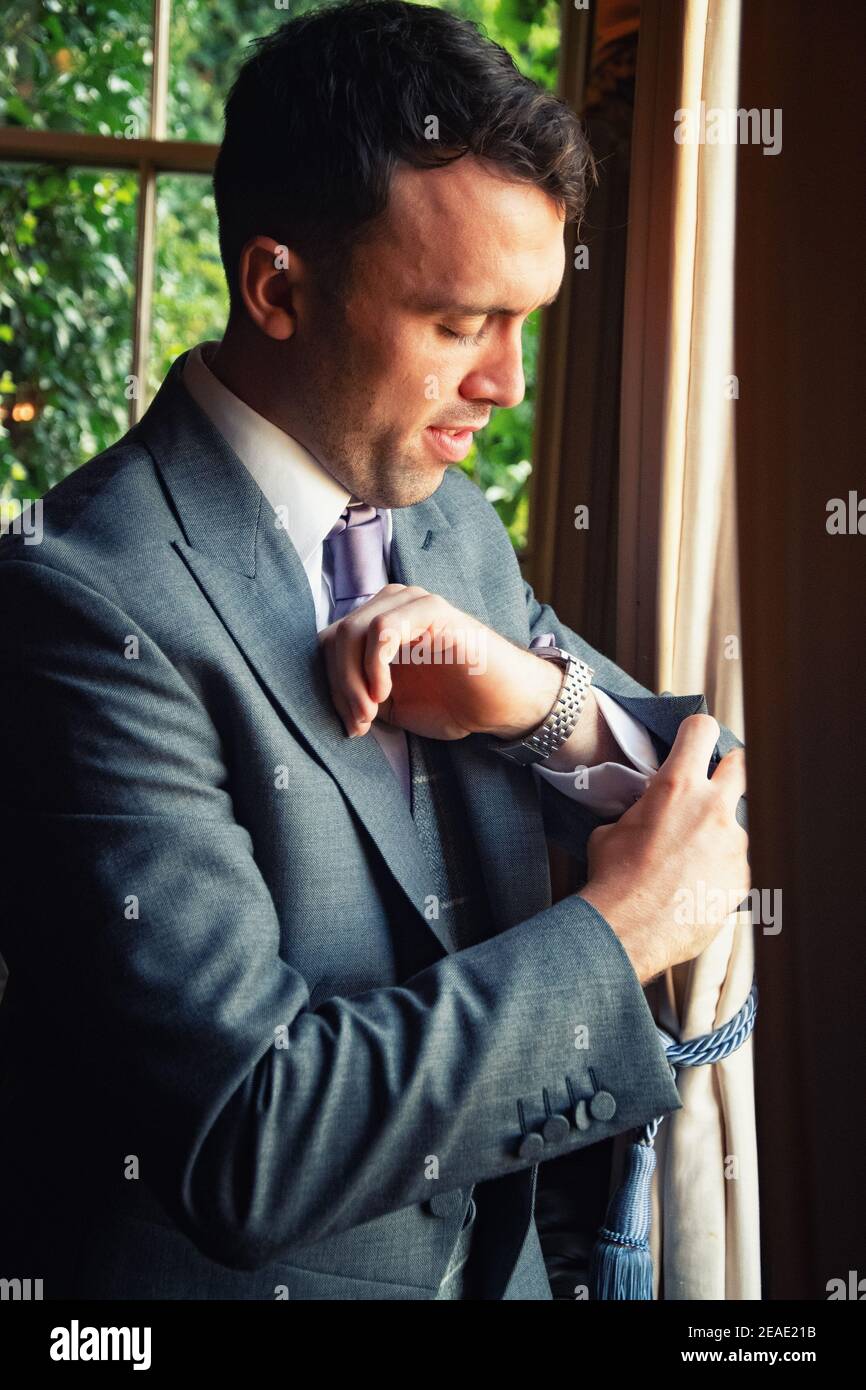 Groom on his wedding day adjusting his cuffs, standing by the window ...