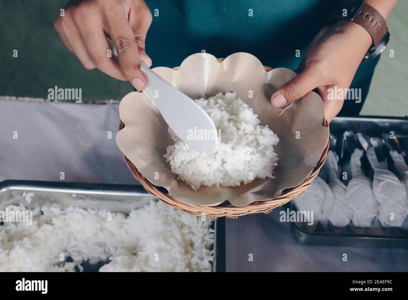 Person hand puts white rice from the steel pan to wooden paper plate ...