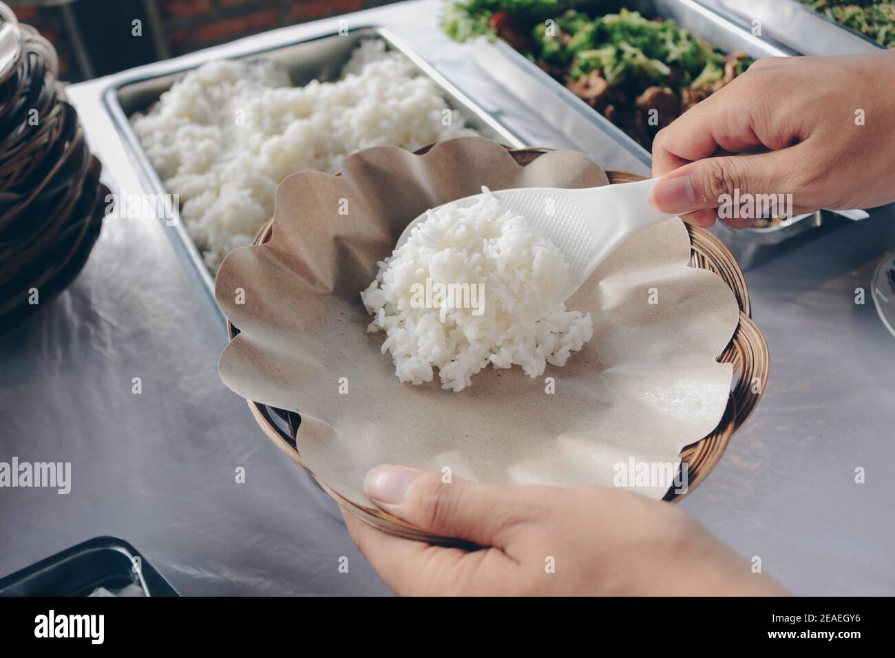 Person hand puts white rice from the steel pan to wooden paper plate ...