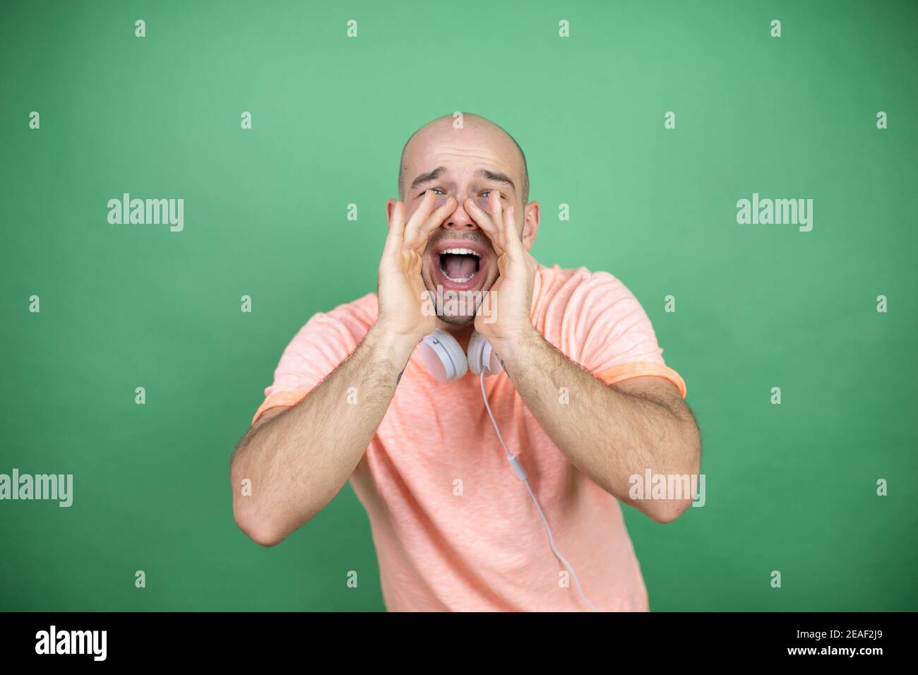Young bald man using headphones over green background shouting and ...