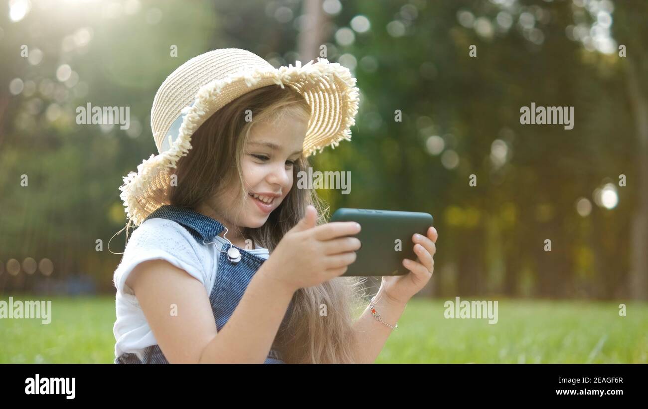 Happy smiling child girl looking in mobile phone outdoors in summer ...