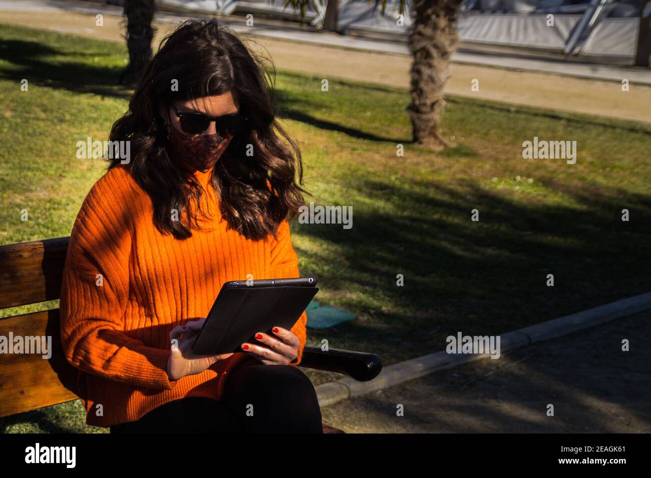 Caucasian female with a face mask sitting on a park bench and using a ...