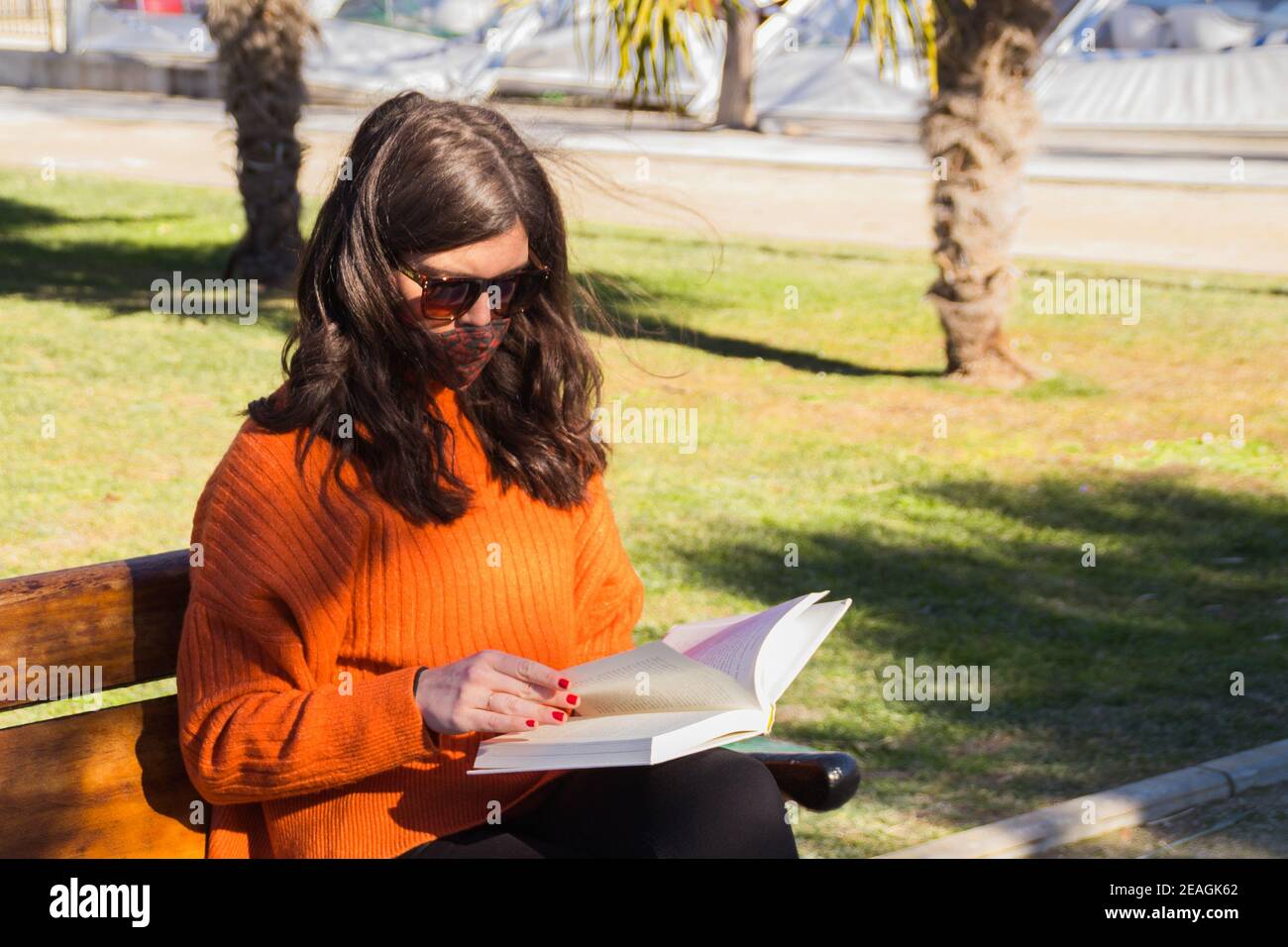 Caucasian female with a face mask and sunglasses sitting on a park ...