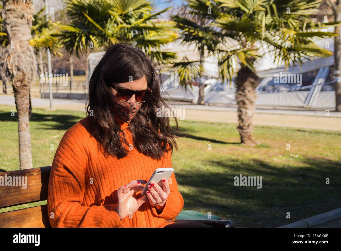 Caucasian female with a face mask sitting on a park bench and using a ...