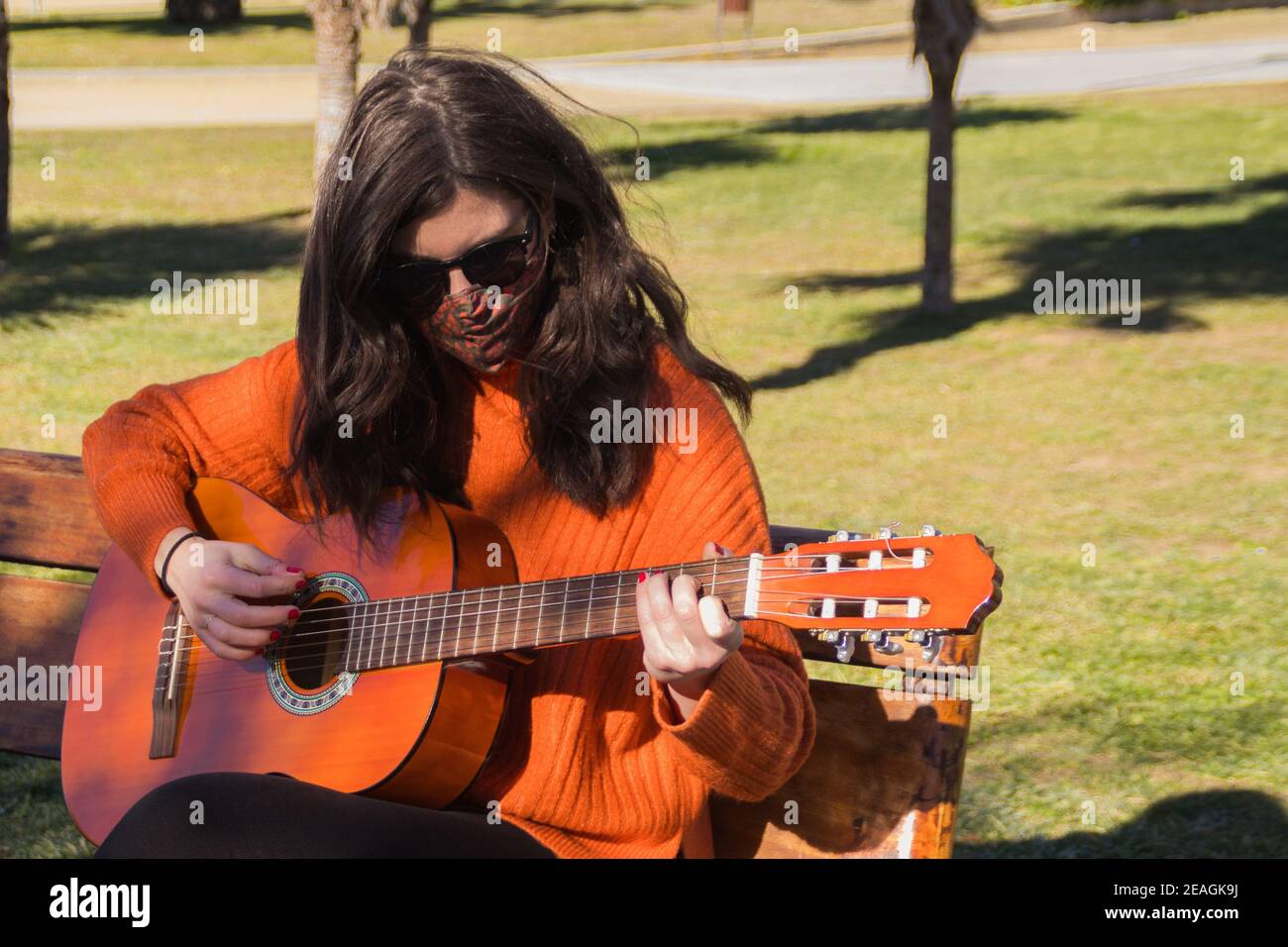 Female with a face mask and sunglasses playing guitar sitting on a park ...