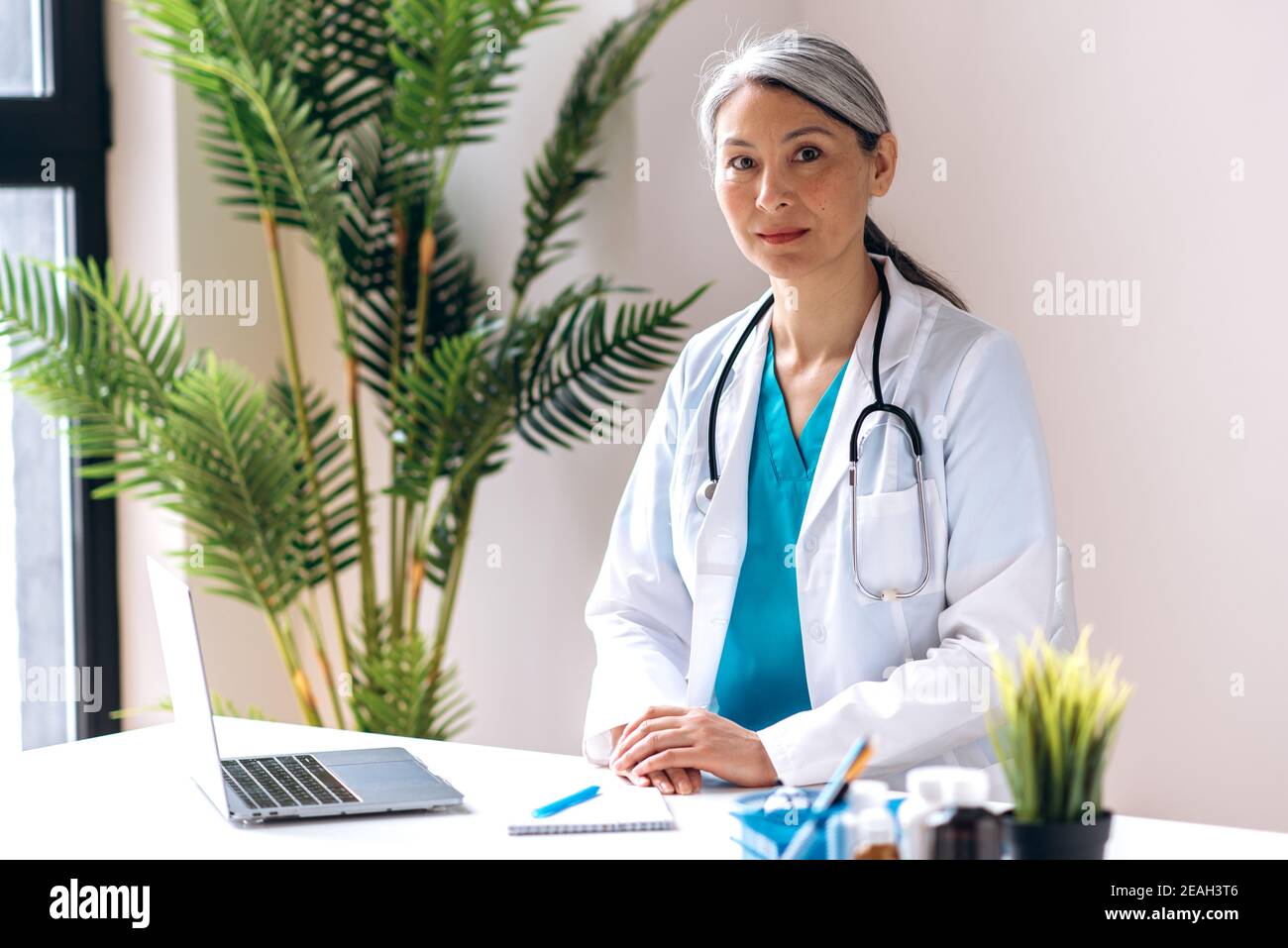 Gray-haired female middle aged general doctor dressed in medical uniform looks directly at the ...