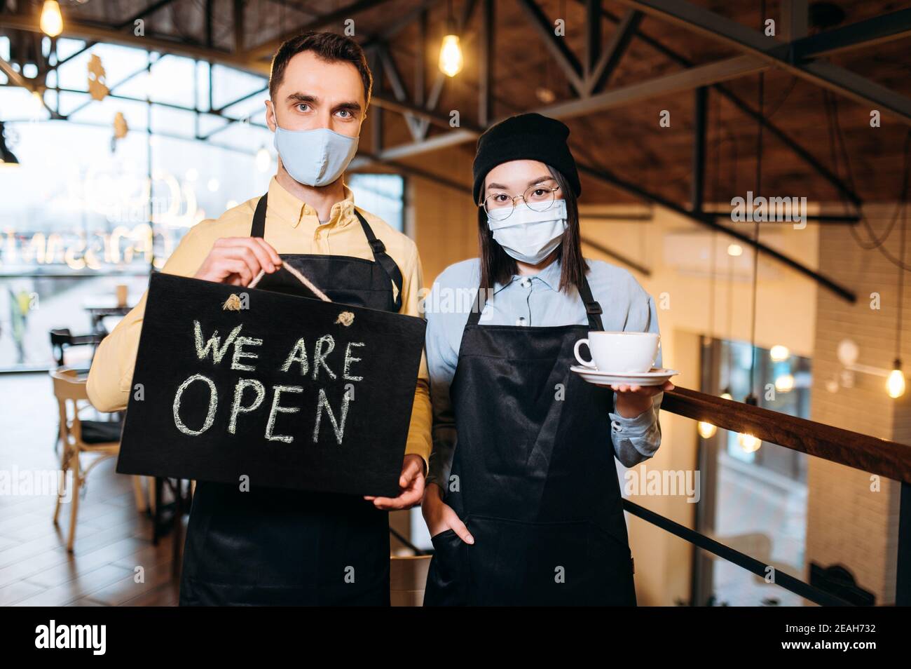 Two friendly multiracial waiters wearing protective masks stand inside ...
