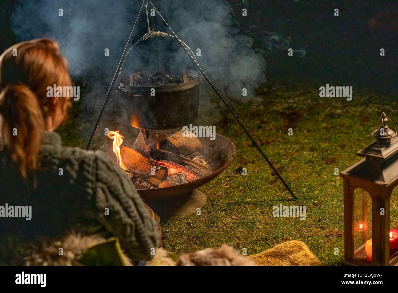 Young blond woman sits outside by the campfire and latern in the woods ...