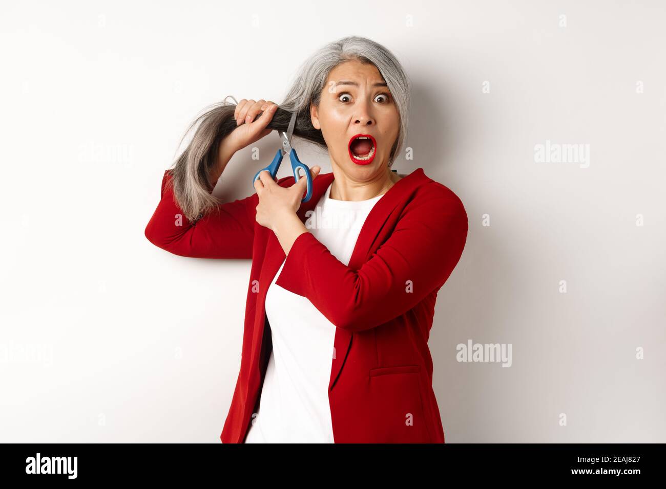 Scared asian senior woman cutting hair with scissors and screaming in ...