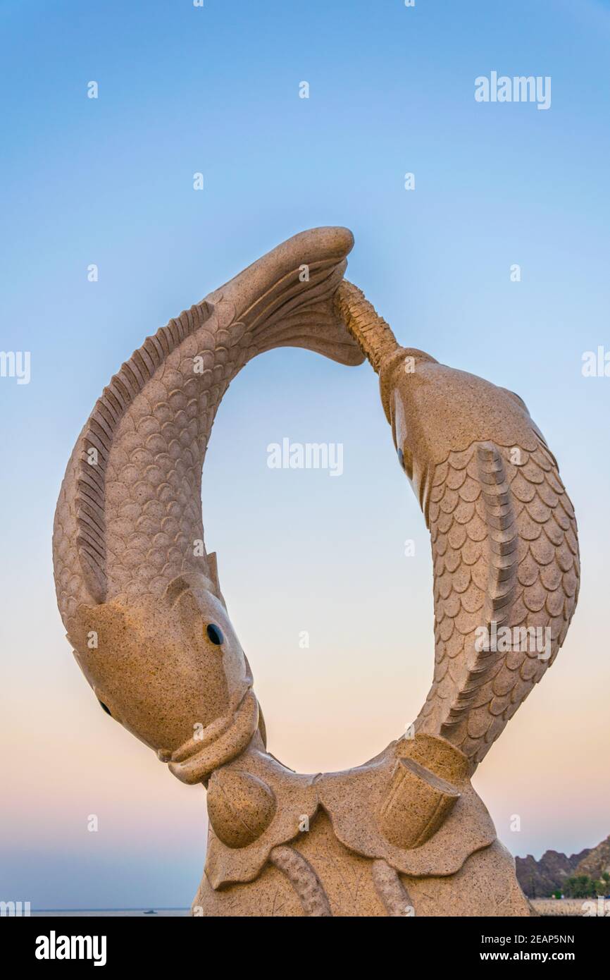 Statue of two fishes on a Muttrah corniche with a fortress on ...