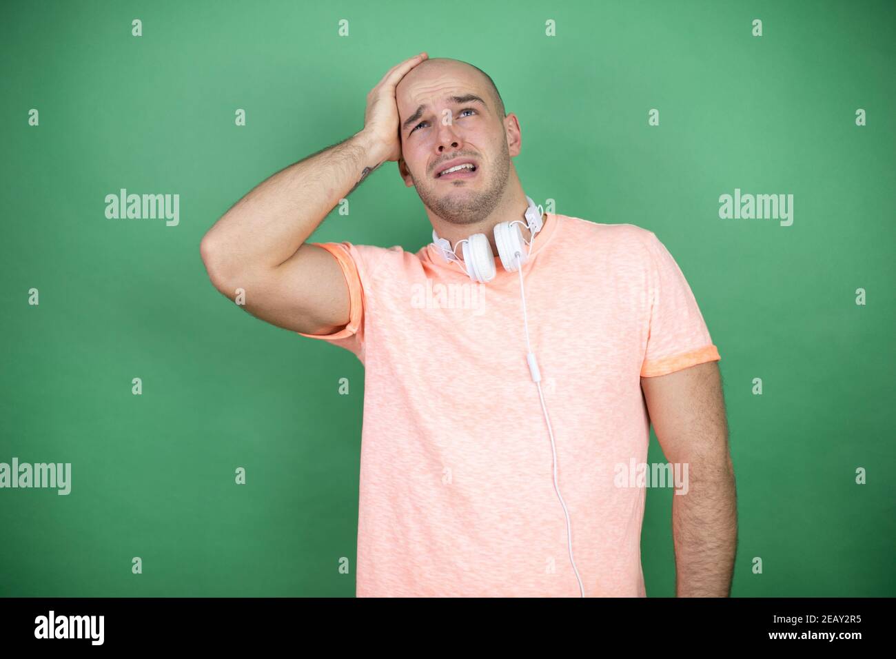 Young bald man using headphones over green background putting one hand ...