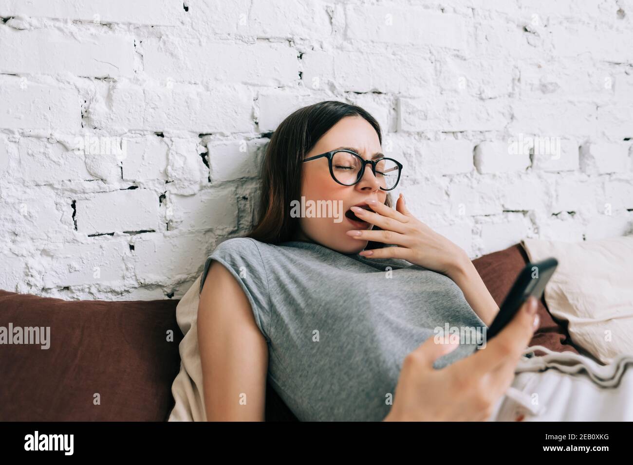Yawning sleepy caucasian woman in eyeglasses using mobile phone while ...