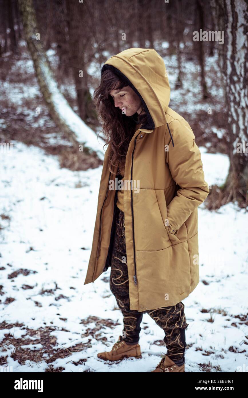 young woman with freckles & long curly hair smiles under hood in snow ...