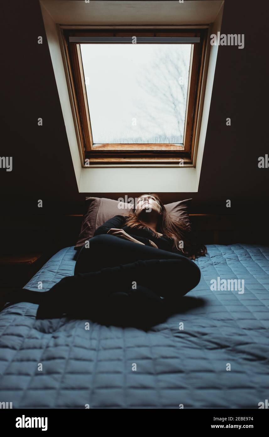 Woman laying on bed in a dark room looking up through a sky light Stock ...