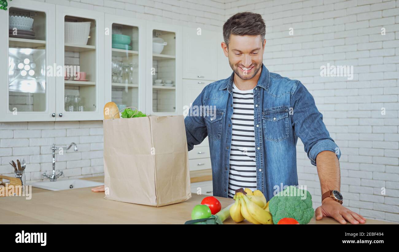 Smiling man looking at fresh food near paper package on kitchen table ...