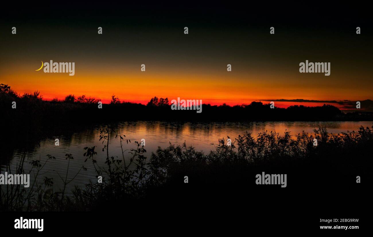 Orange-red sunset sky over a river with silhouetted grass and trees ...