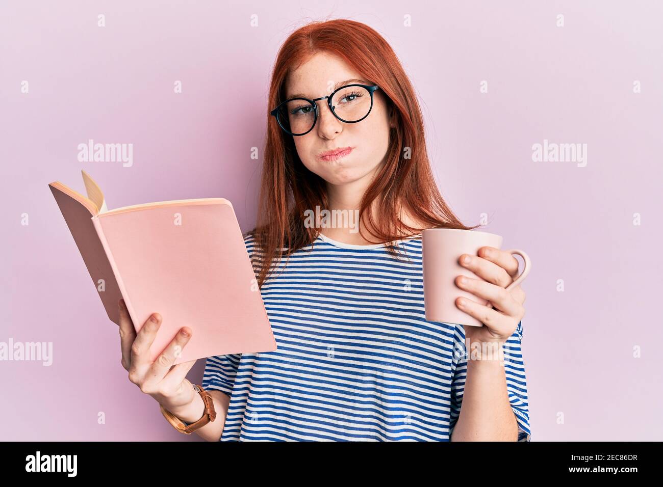 Young red head girl reading a book and drinking a cup of coffee puffing cheeks with funny face ...