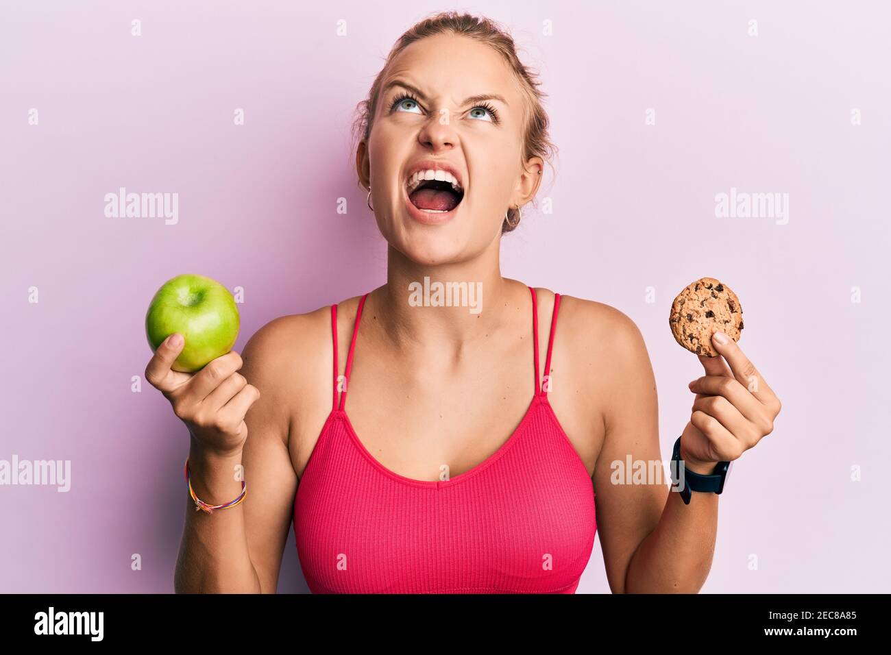 Beautiful caucasian woman holding green apple and cookie angry and mad ...