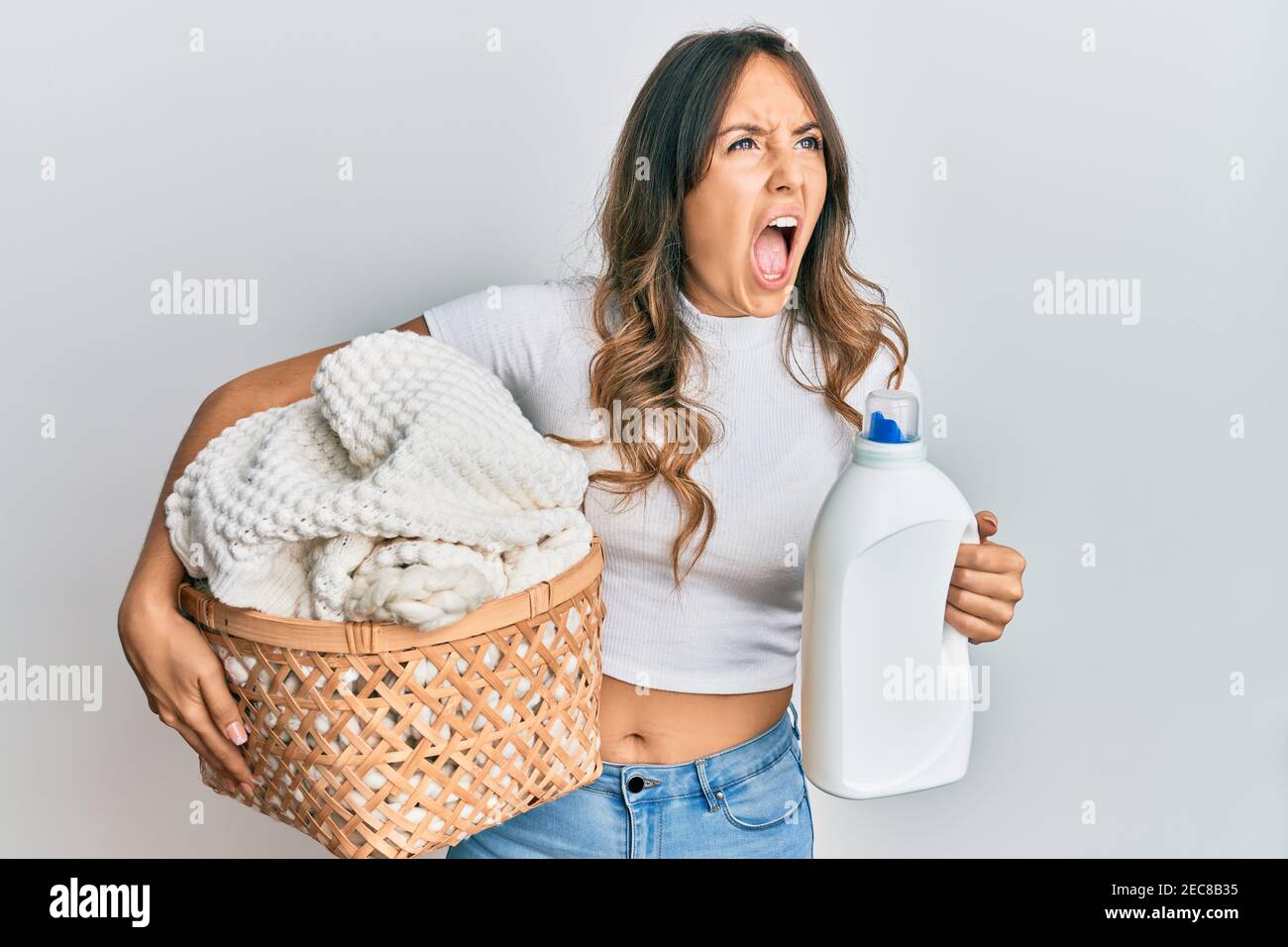 Young brunette woman holding laundry basket and detergent bottle angry and mad screaming ...