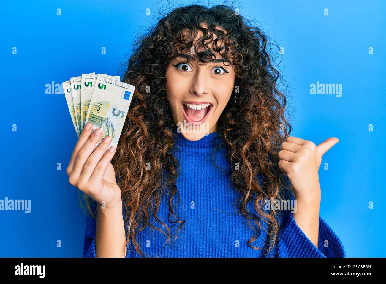 Young hispanic girl holding euro banknotes pointing thumb up to the ...