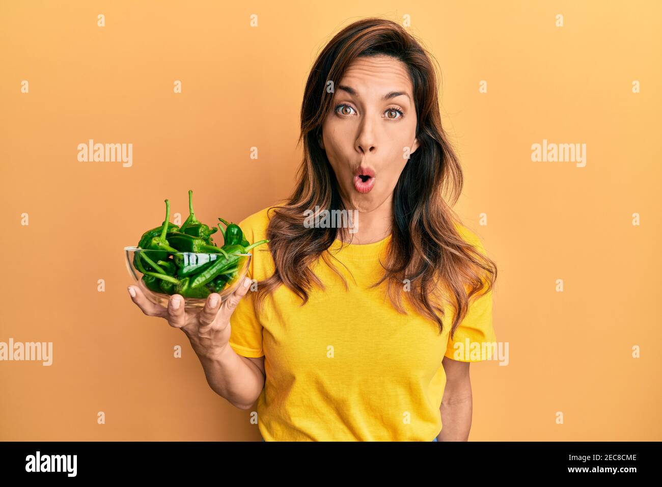 Young latin woman holding bowl with green peppers scared and amazed ...