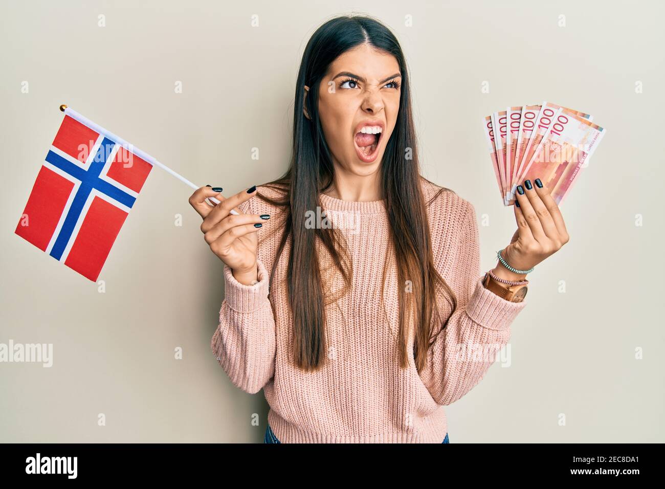 Young hispanic woman holding norway flag and krone banknotes angry and ...