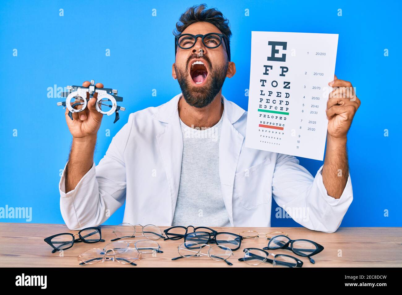 Young hispanic man with optometry glasses and paper with letters angry ...