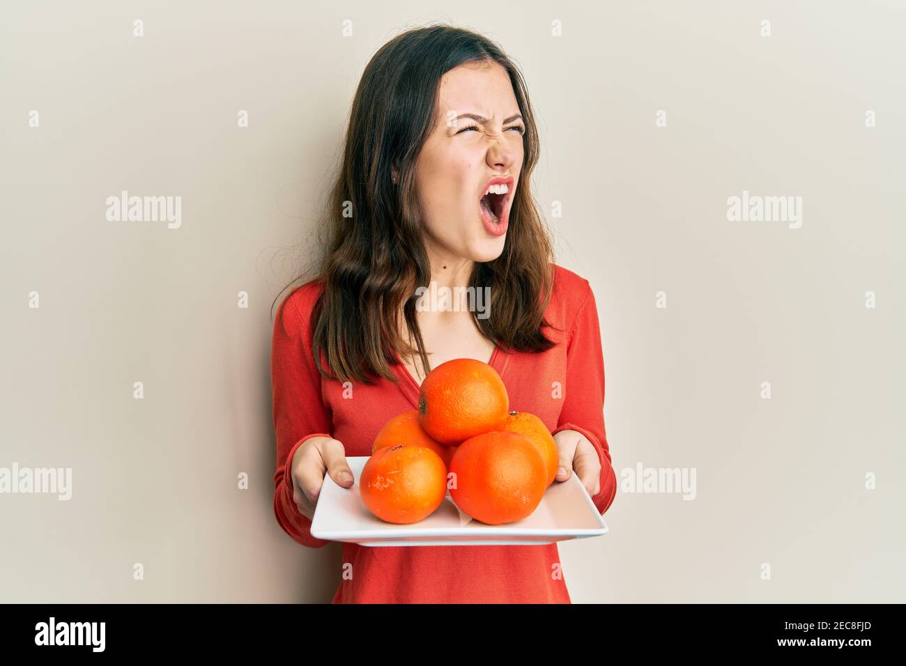 Young brunette woman holding plate with fresh oranges angry and mad ...
