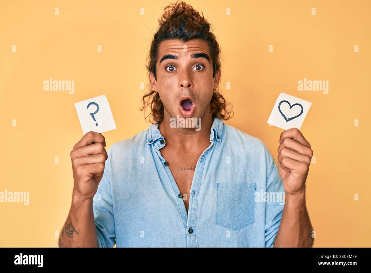 Young hispanic man holding heart and question mark reminder afraid and ...