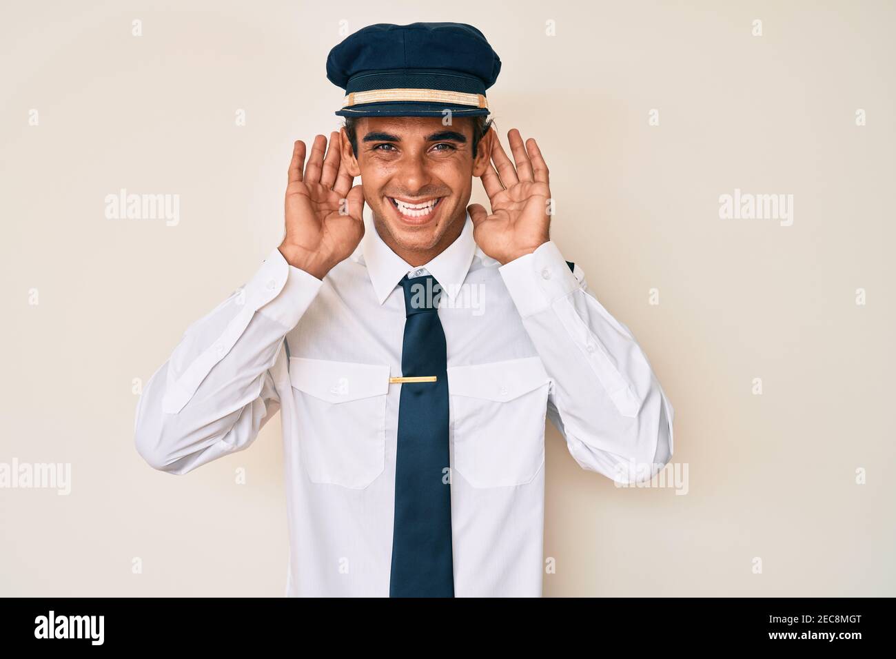 Young hispanic man wearing airplane pilot uniform trying to hear both ...