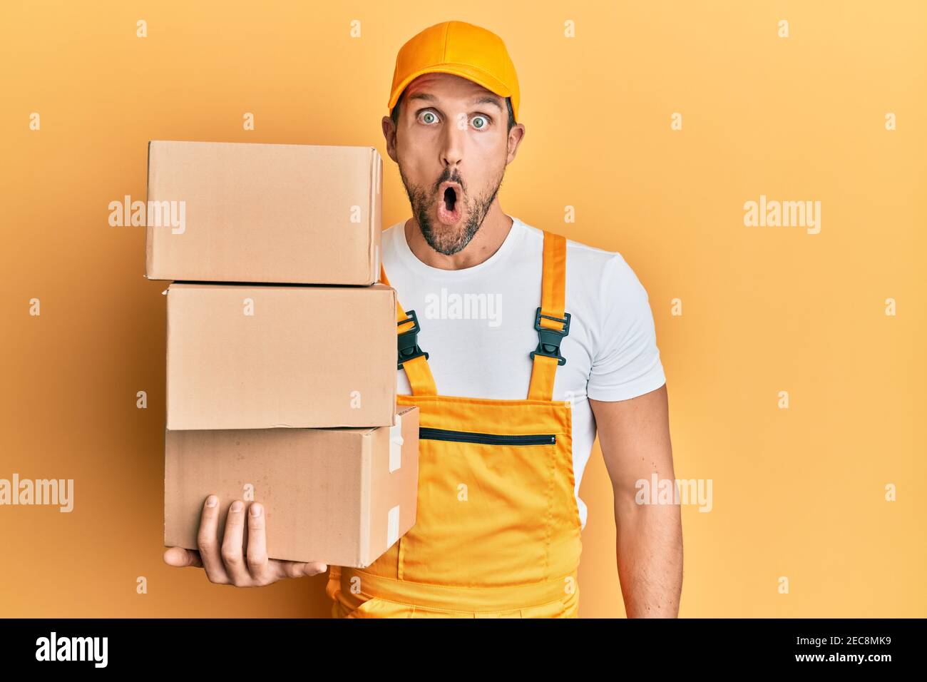 Young handsome delivery man holding packages scared and amazed with ...