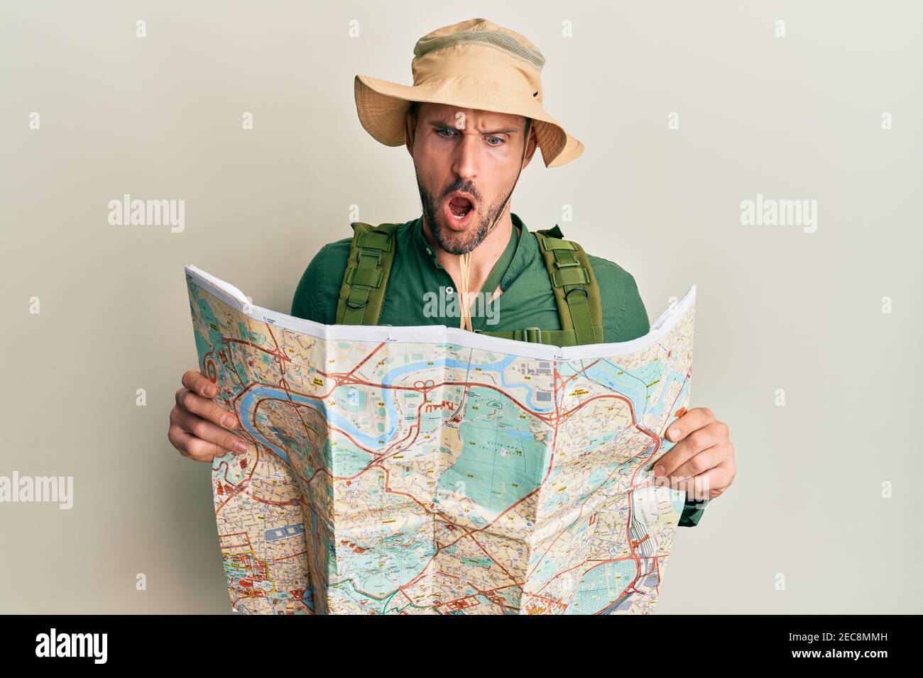 Handsome man with beard wearing explorer hat holding map in shock face ...