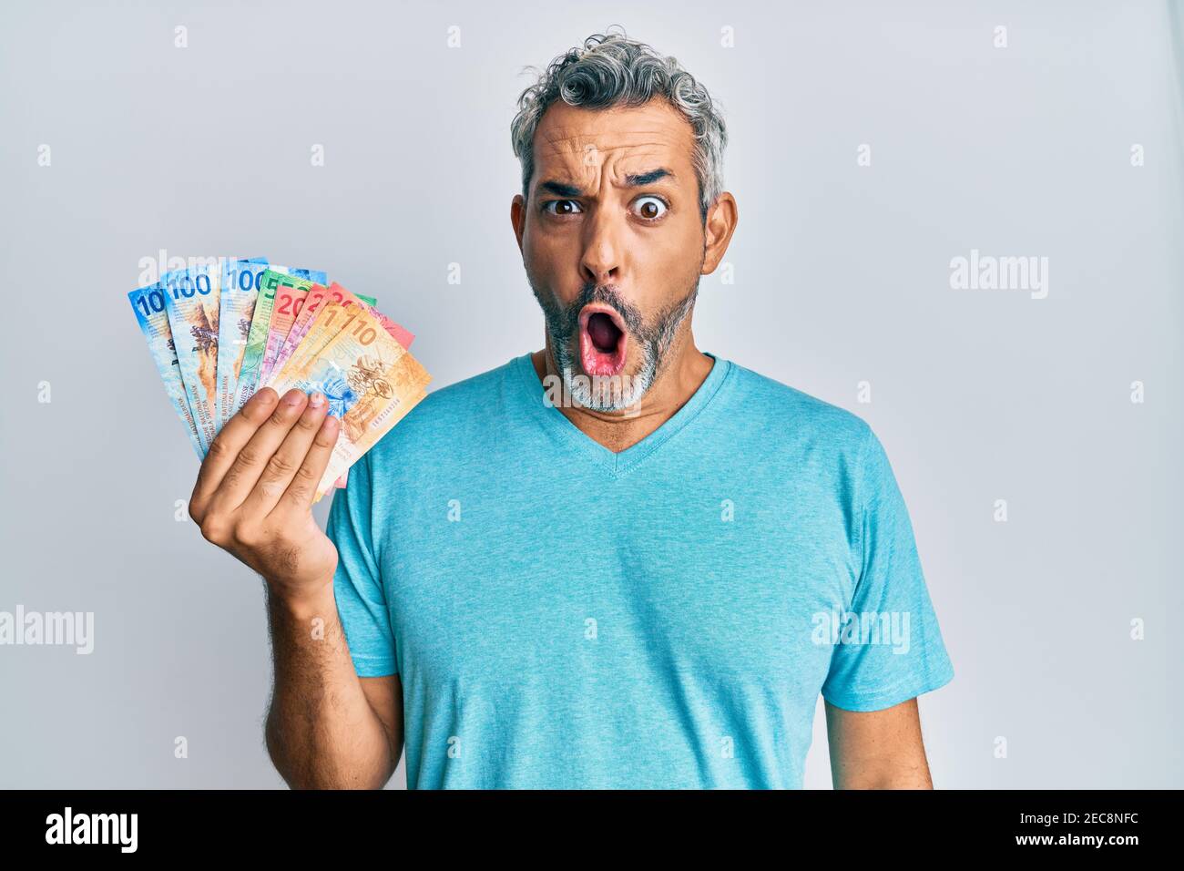 Middle age grey-haired man holding swiss franc banknotes scared and ...