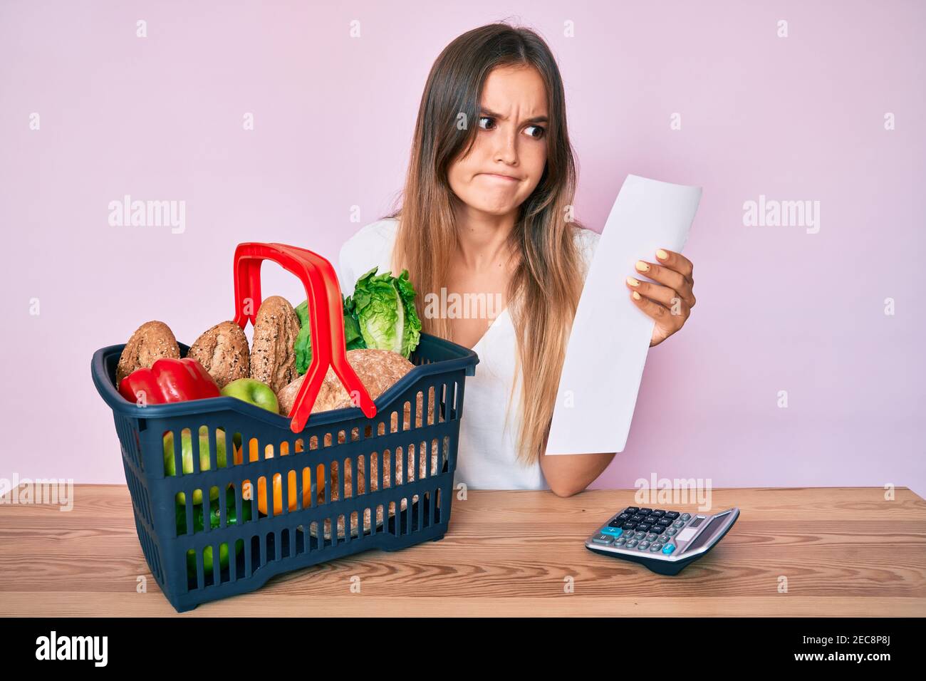 Beautiful caucasian woman holding supermarket basket and groceries list ...
