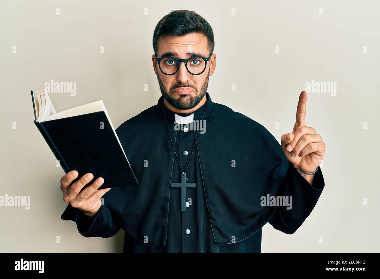 Young hispanic priest man holding bible with finger up depressed and ...