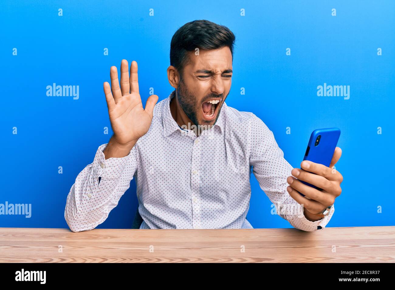 Handsome hispanic man doing video call waving to smartphone angry and ...