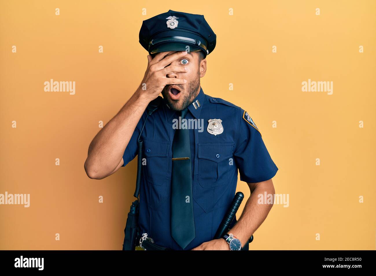 Handsome hispanic man wearing police uniform peeking in shock covering ...