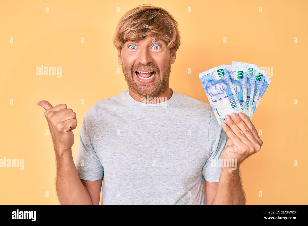 Young blond man holding south african rand banknotes pointing thumb up ...