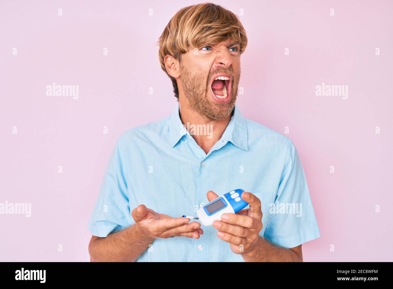 Young blond man using glucose meter angry and mad screaming frustrated ...