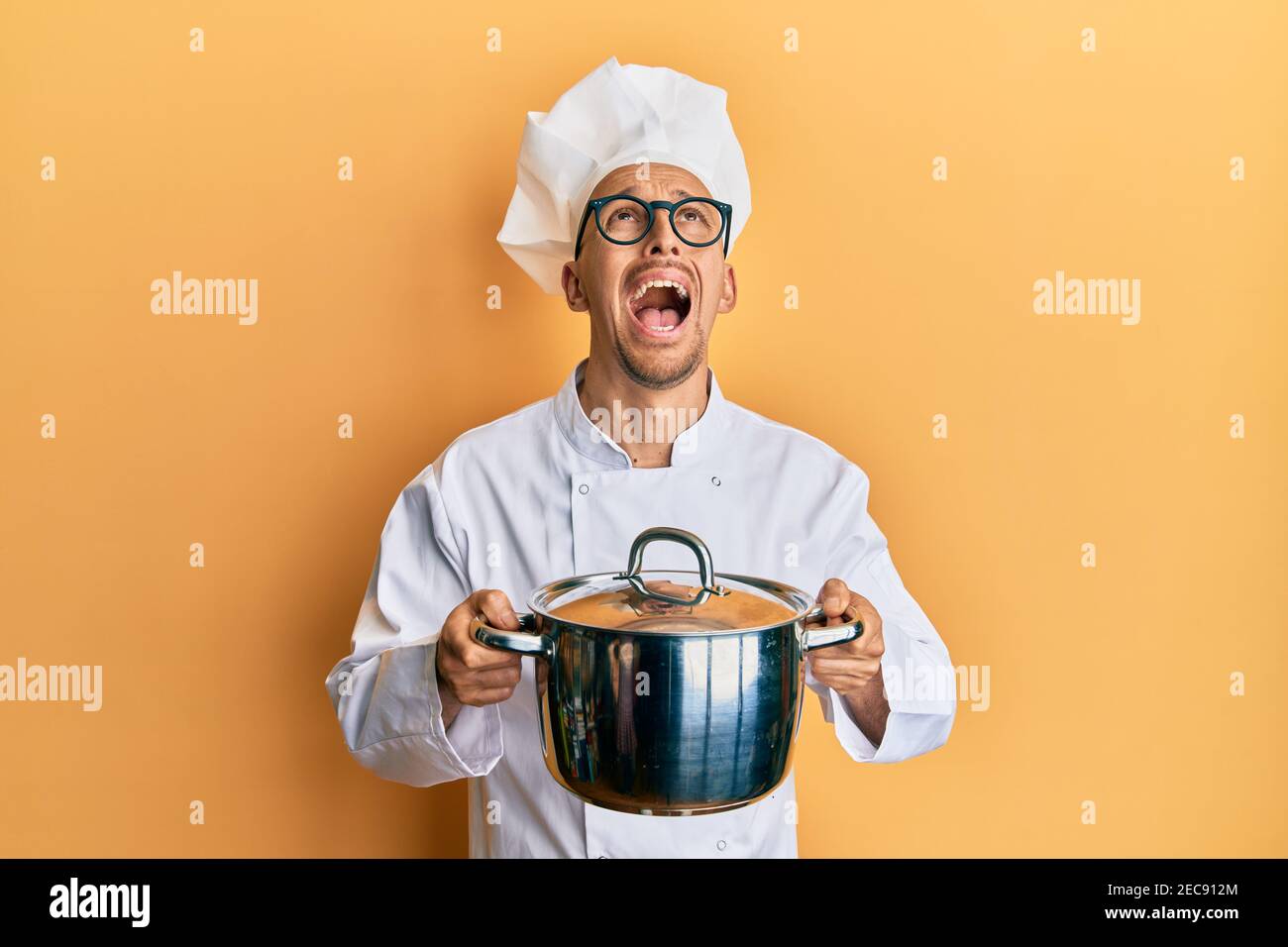 Bald man with beard wearing professional cook holding cooking pot angry ...