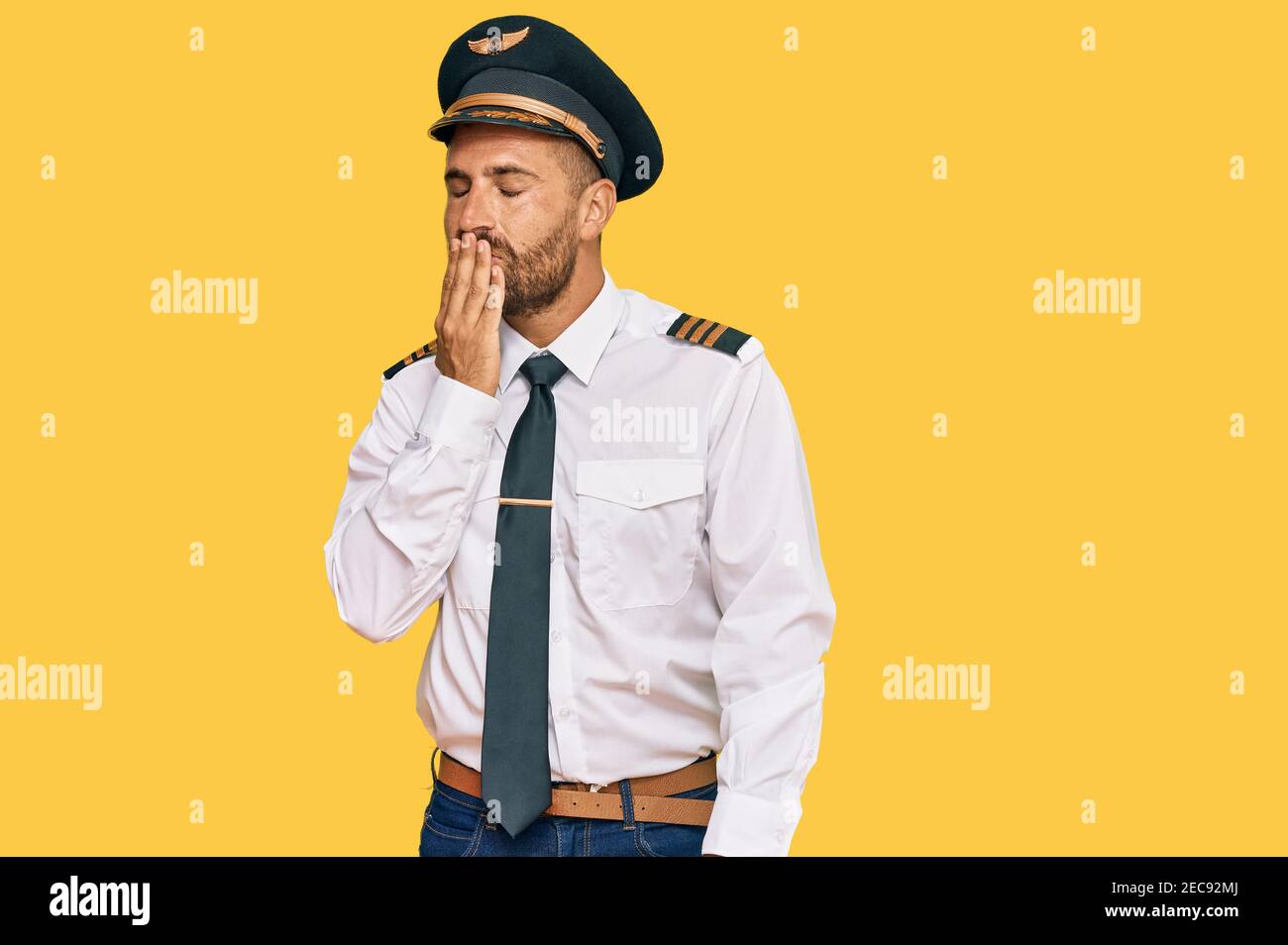 Handsome man with beard wearing airplane pilot uniform bored yawning ...