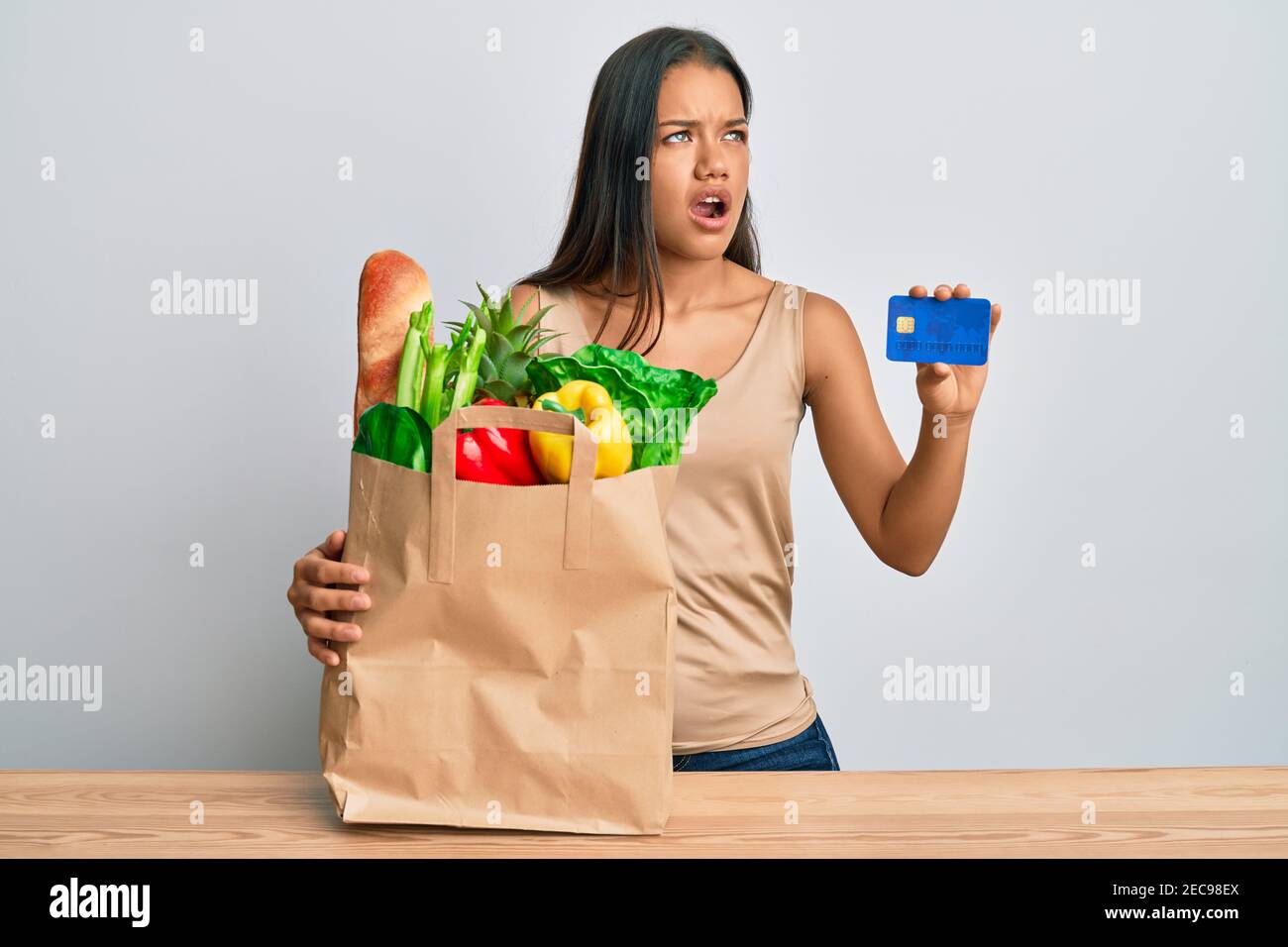 Beautiful hispanic woman holding groceries and credit card angry and ...