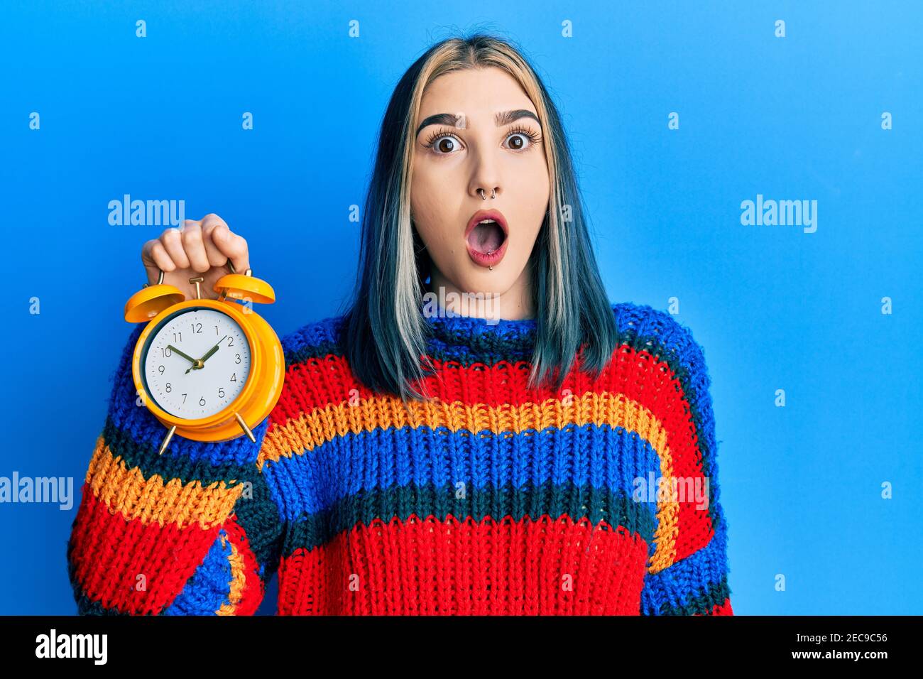 Young modern girl holding alarm clock scared and amazed with open mouth ...