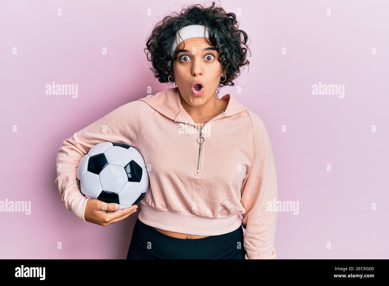 Young hispanic woman with curly hair holding soccer ball scared and ...