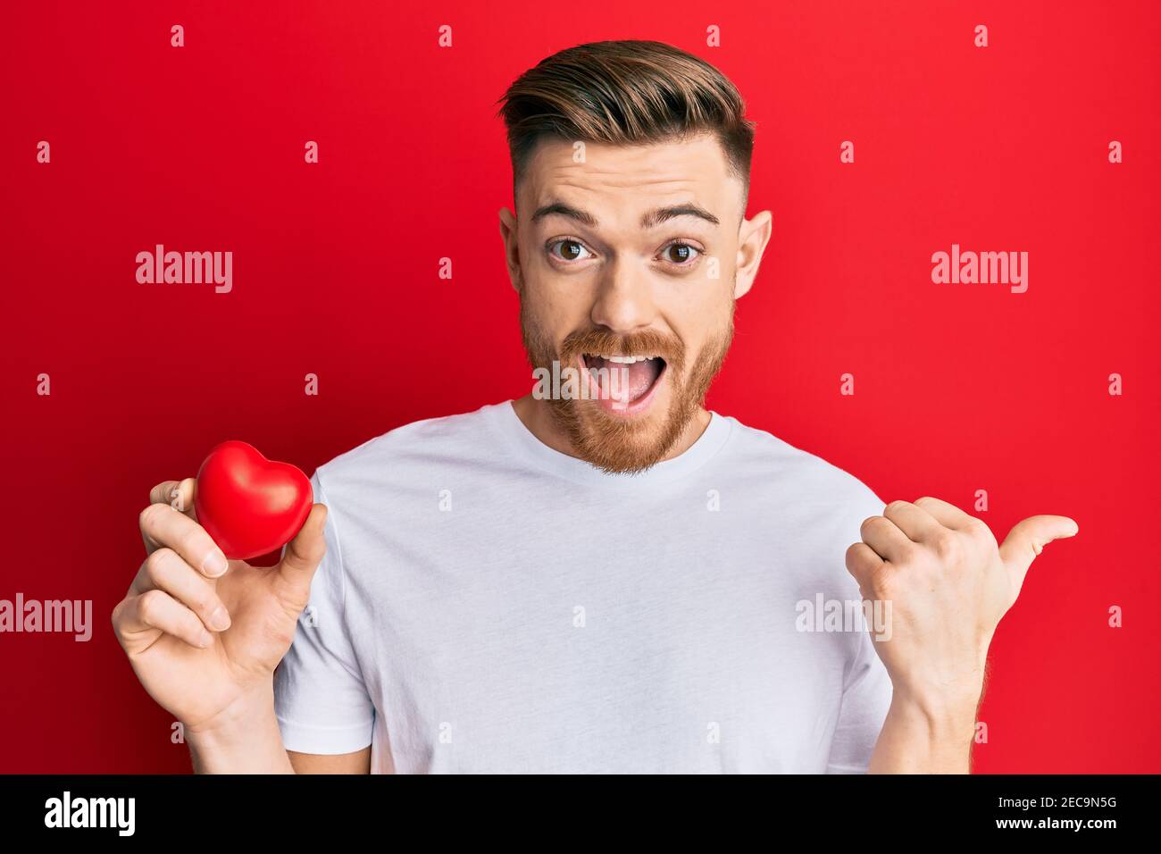 Young redhead man holding heart pointing thumb up to the side smiling ...