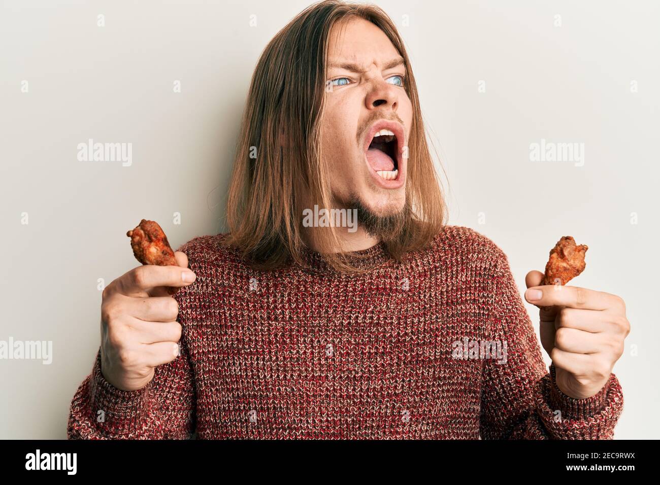 Handsome caucasian man with long hair eating chicken wings angry and ...